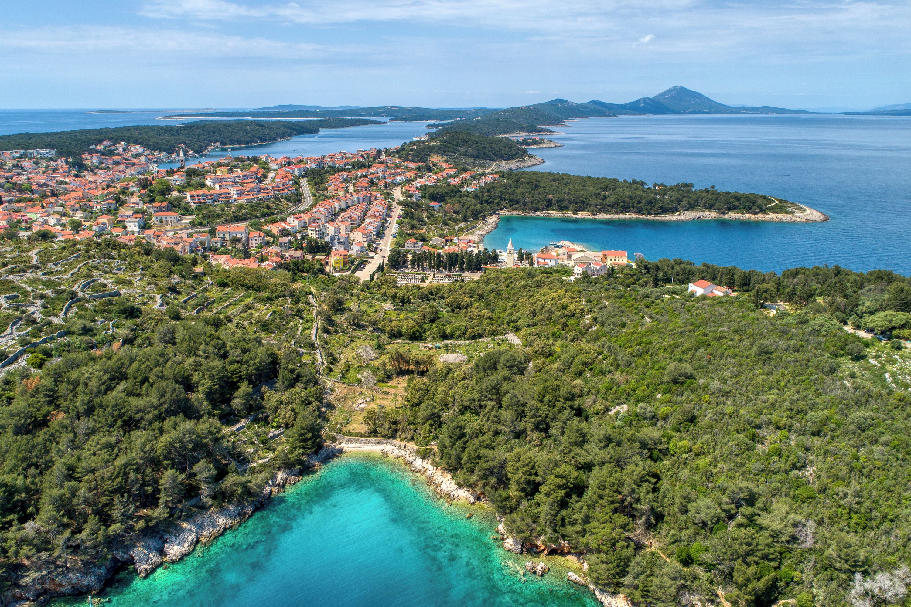 Vista aérea de uma vila costeira com uma baía turquesa translúcida, áreas florestais verdes e densas, e uma igreja imponente.