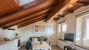 Cozinha moderna com armários brancos, área de jantar com mesa e cadeiras, TV fixada na parede e teto de madeira.