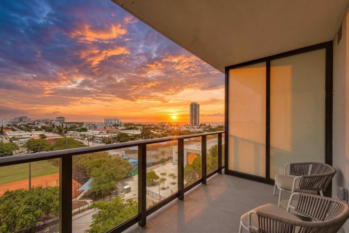 Varanda com vista para o pôr do sol sobre uma paisagem urbana, incluindo um edifício alto e um campo esportivo.