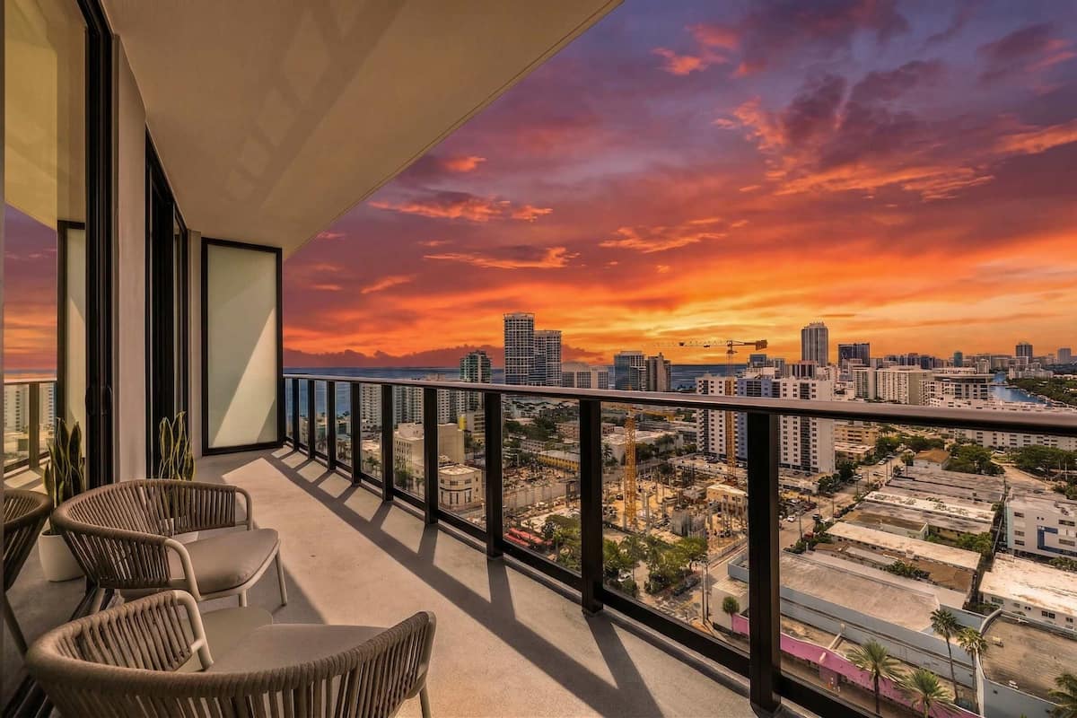 Varanda com móveis de vime, vista para a cidade e céu ao pôr do sol.