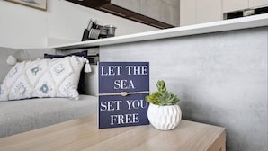 Uma sala de estar com sofá, mesa de centro, um letreiro com a frase 'LET THE SEA SET YOU FREE' e uma planta em vaso.