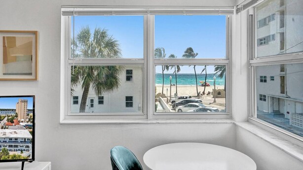 Quarto com uma janela grande de frente para uma praia, uma mesa redonda e uma cadeira.