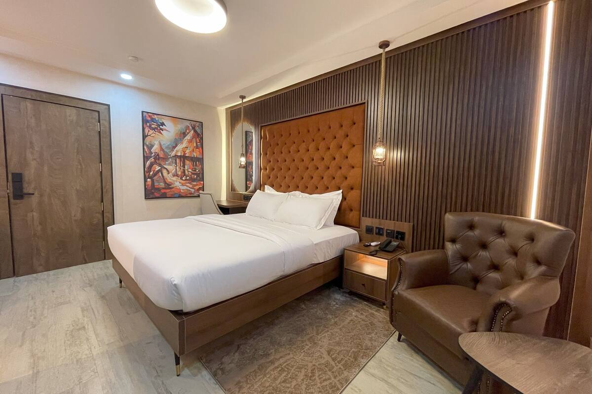 Quarto de hotel com uma cama grande, cabeceira de madeira, mesa de cabeceira, poltrona de couro e um quadro na parede.