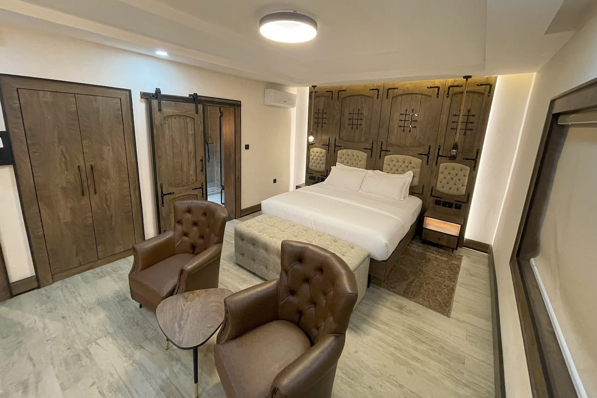Quarto de hotel com uma cama grande, duas poltronas de couro marrom, uma mesinha e um guarda-roupa com espelho.