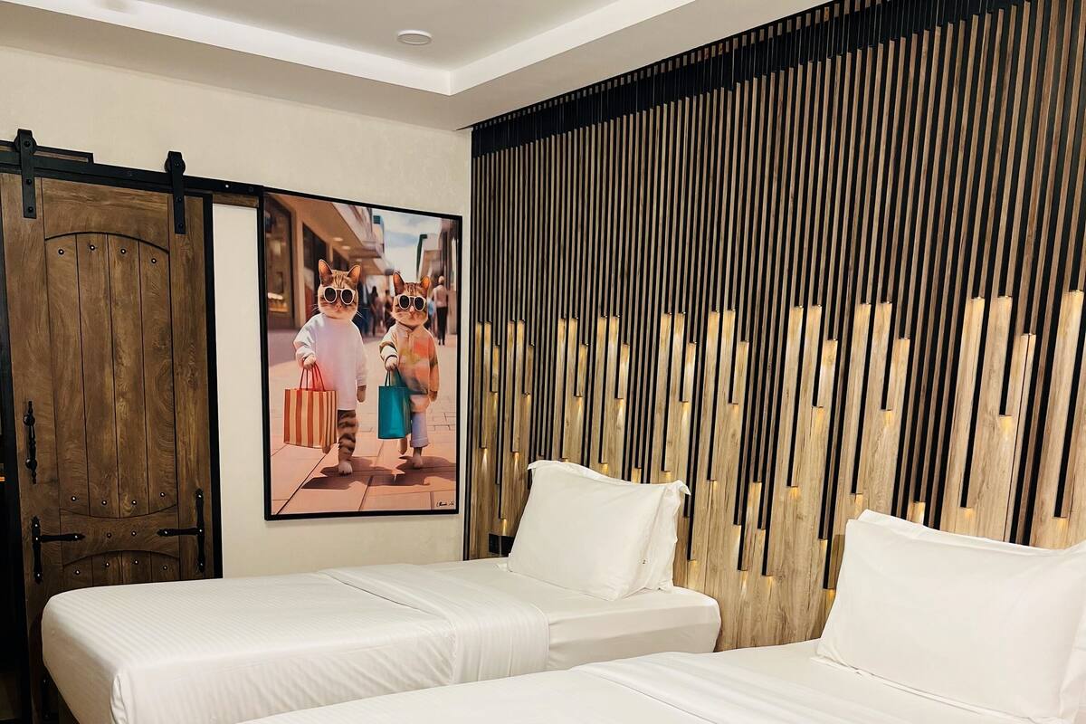 Quarto de hotel moderno com duas camas, uma porta de madeira, um quadro pendurado na parede e um tapete estampado.