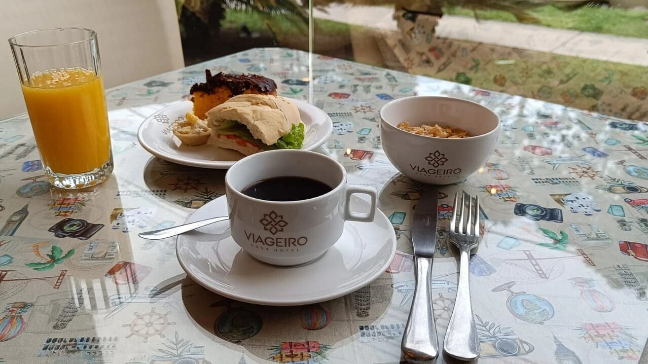 Uma mesa com itens de café da manhã, incluindo um copo de suco de laranja, um sanduíche, duas xícaras de café e uma tigela de cereal.
