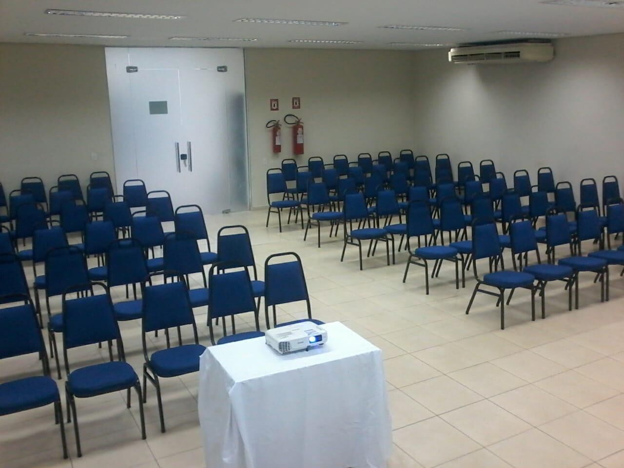 Uma sala de conferências com fileiras de cadeiras azuis, uma mesa branca com projetor e uma porta com extintor na parede.