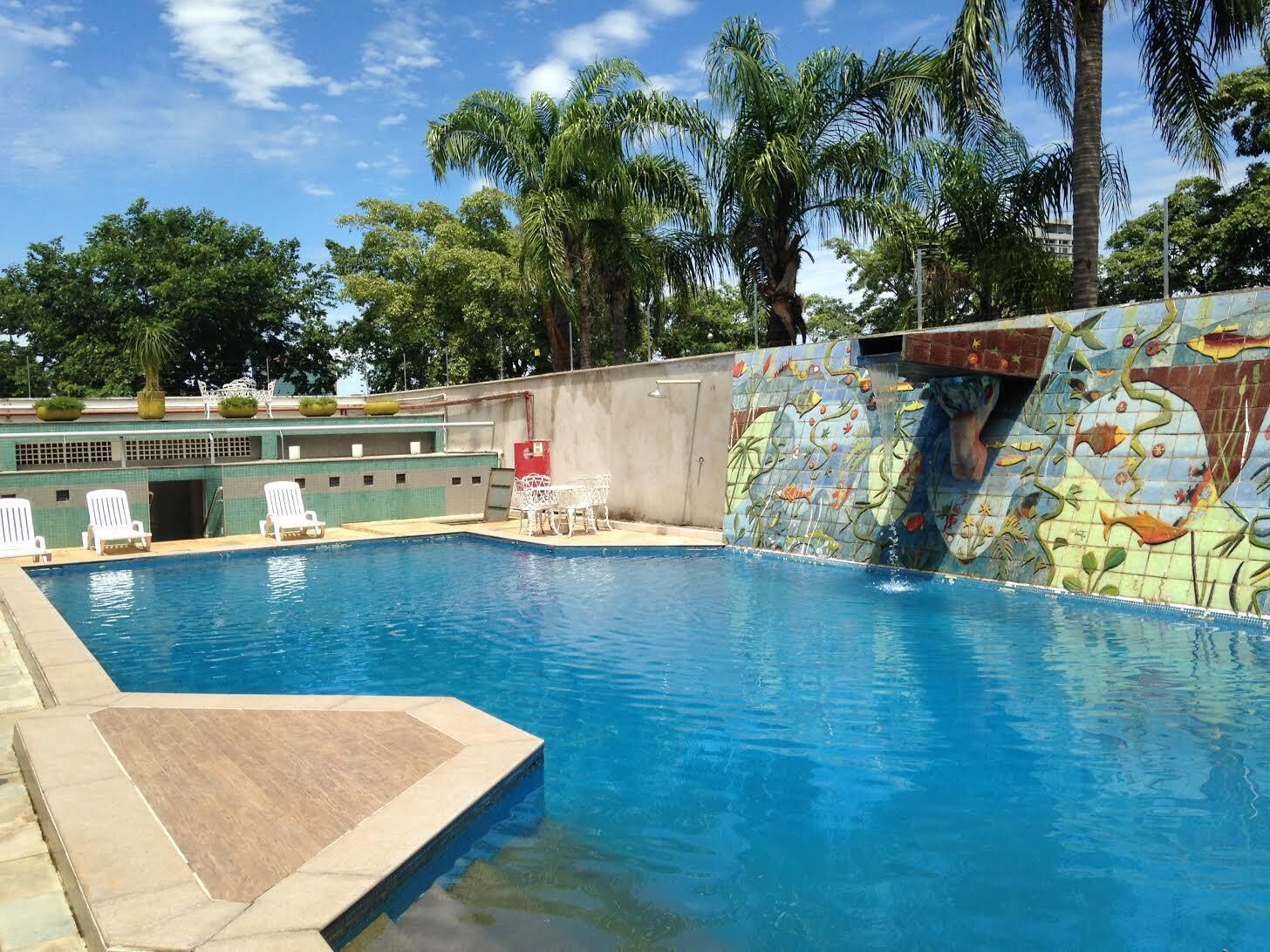 Piscina com água azul cristalina, rodeada por palmeiras e uma parede revestida com um mural colorido.