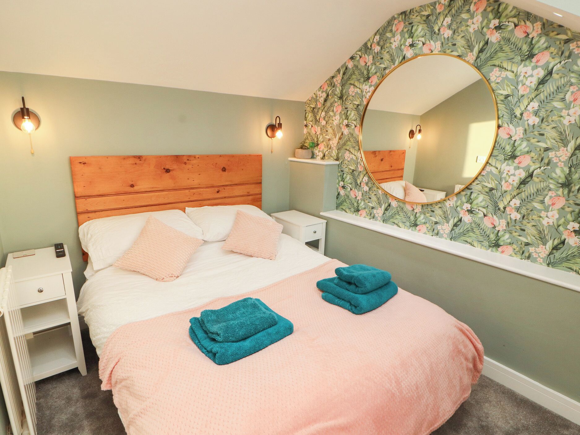 Quarto com cabeceira de madeira, cama com roupa de cama rosa, duas toalhas verde-água, mesa de cabeceira, gaveta pequena, espelho fixado na parede e papel de parede floral.