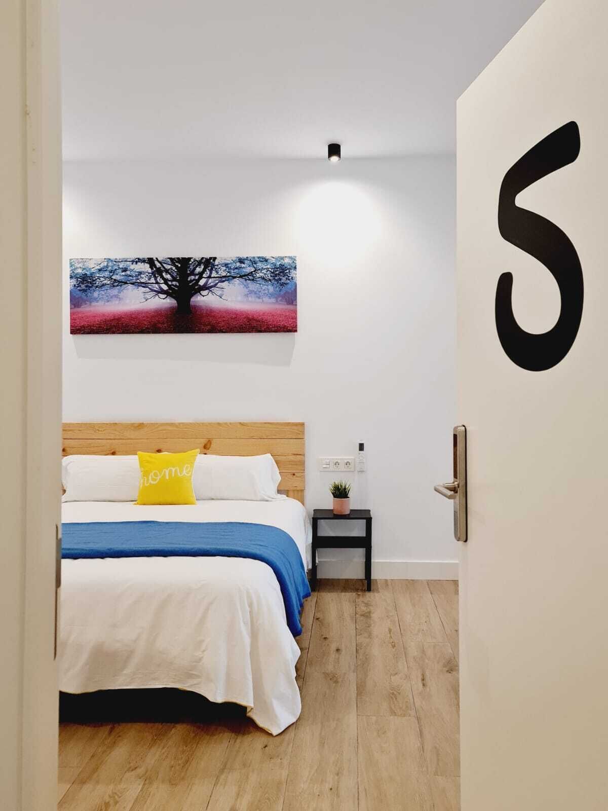 Um quarto com cama, mesa de cabeceira, um quadro na parede e uma porta com um número grande.