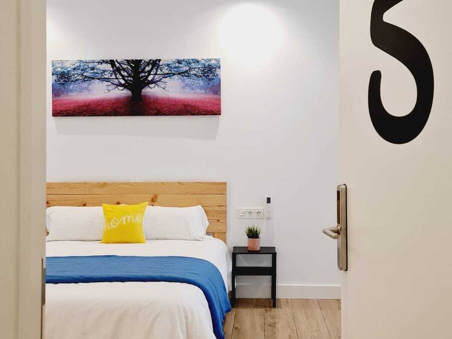 Um quarto com cama, mesa de cabeceira, um quadro na parede e uma porta com um número grande.