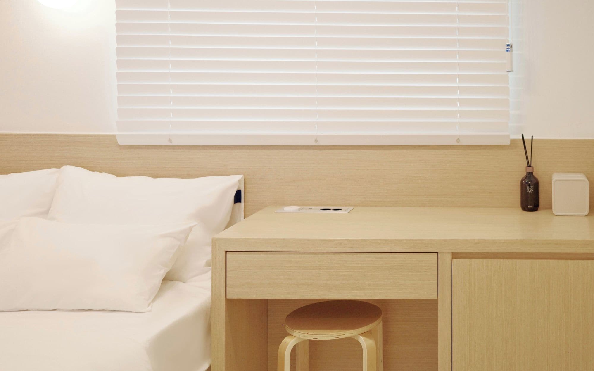 Quarto de hotel com cama, escrivaninha e cadeira, janela com persianas, e uma mesinha com garrafa e recipiente.