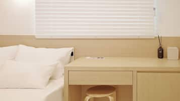 Quarto de hotel com cama, escrivaninha e cadeira, janela com persianas, e uma mesinha com garrafa e recipiente.