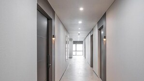 Um corredor comprido e estreito, com portas em ambos os lados, luzes embutidas no teto e piso de azulejos.