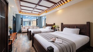 Quarto de hotel com duas camas, um sofá, um banco de madeira e uma janela grande com vista para edifícios.