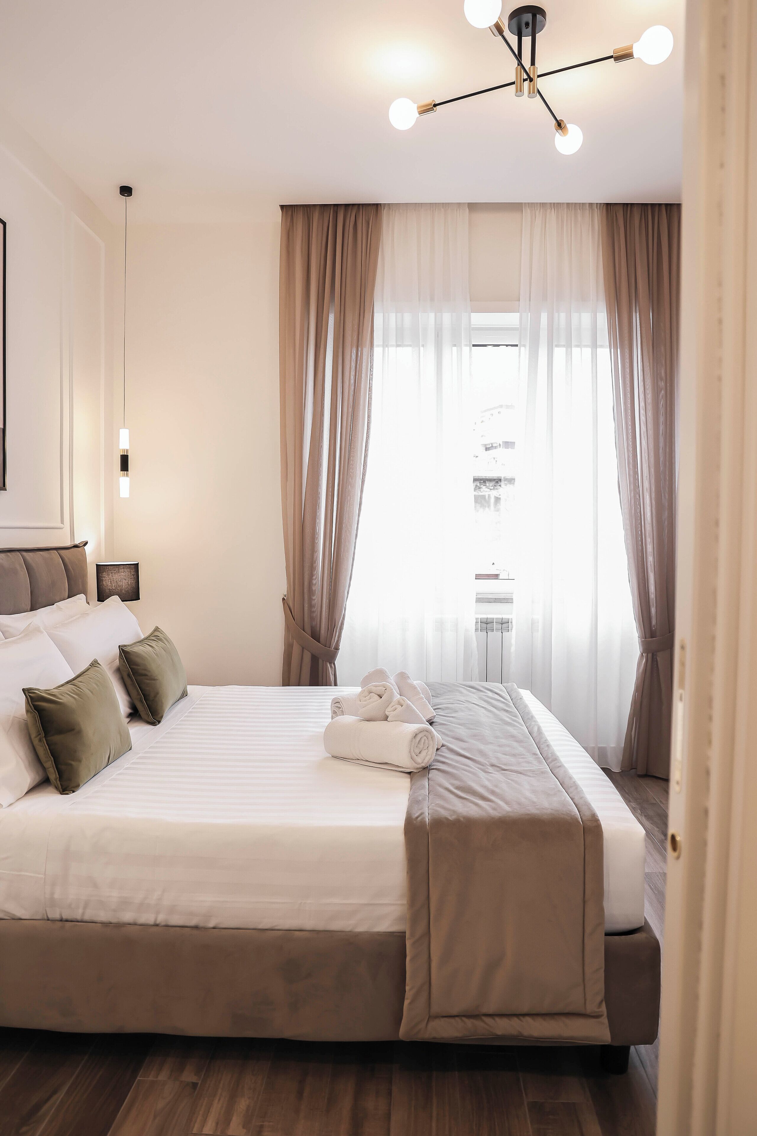 Cama bem arrumada com colcha bege e almofadas, um lustre moderno e cortinas leves.