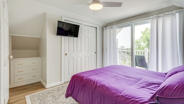 Quarto com cama, televisão, ventilador de teto, porta de vidro deslizante e uma pequena varanda.