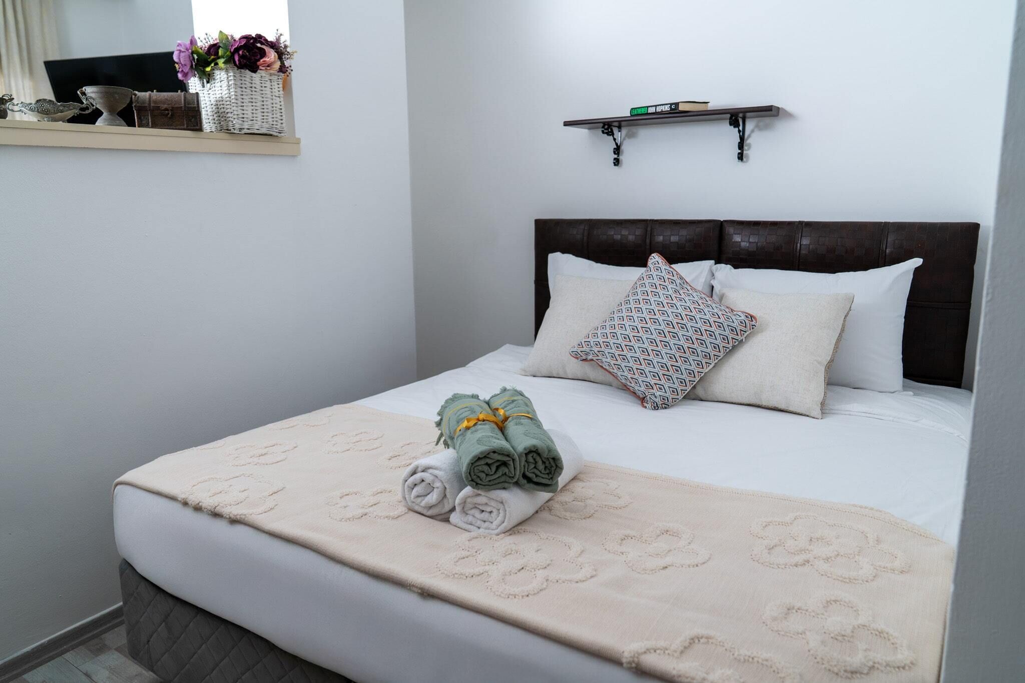 Cama bem arrumada com travesseiro estampado, toalha enrolada e cesta decorativa numa prateleira acima.