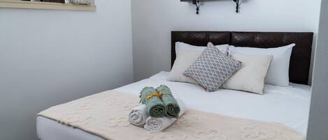 Cama bem arrumada com travesseiro estampado, toalha enrolada e cesta decorativa numa prateleira acima.