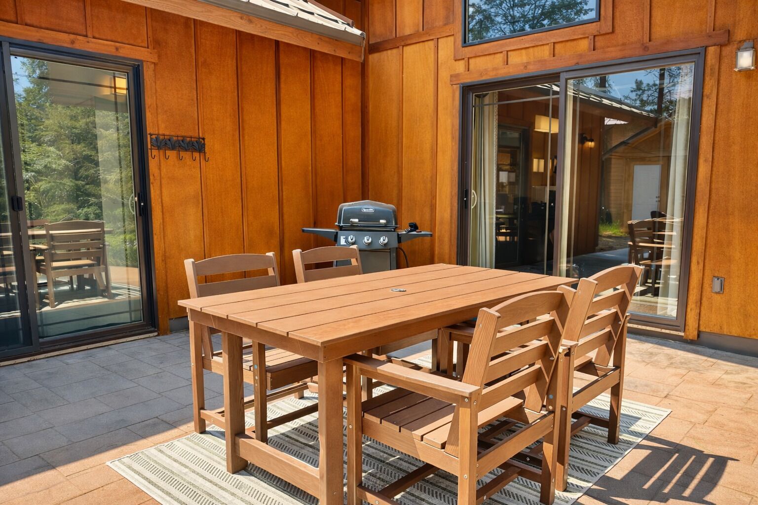 Um pátio externo com mesa e cadeiras de madeira, churrasqueira e portas de vidro deslizantes que dão para um espaço interno.
