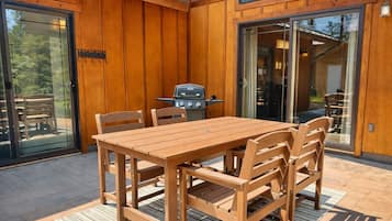 Um pátio externo com mesa e cadeiras de madeira, churrasqueira e portas de vidro deslizantes que dão para um espaço interno.
