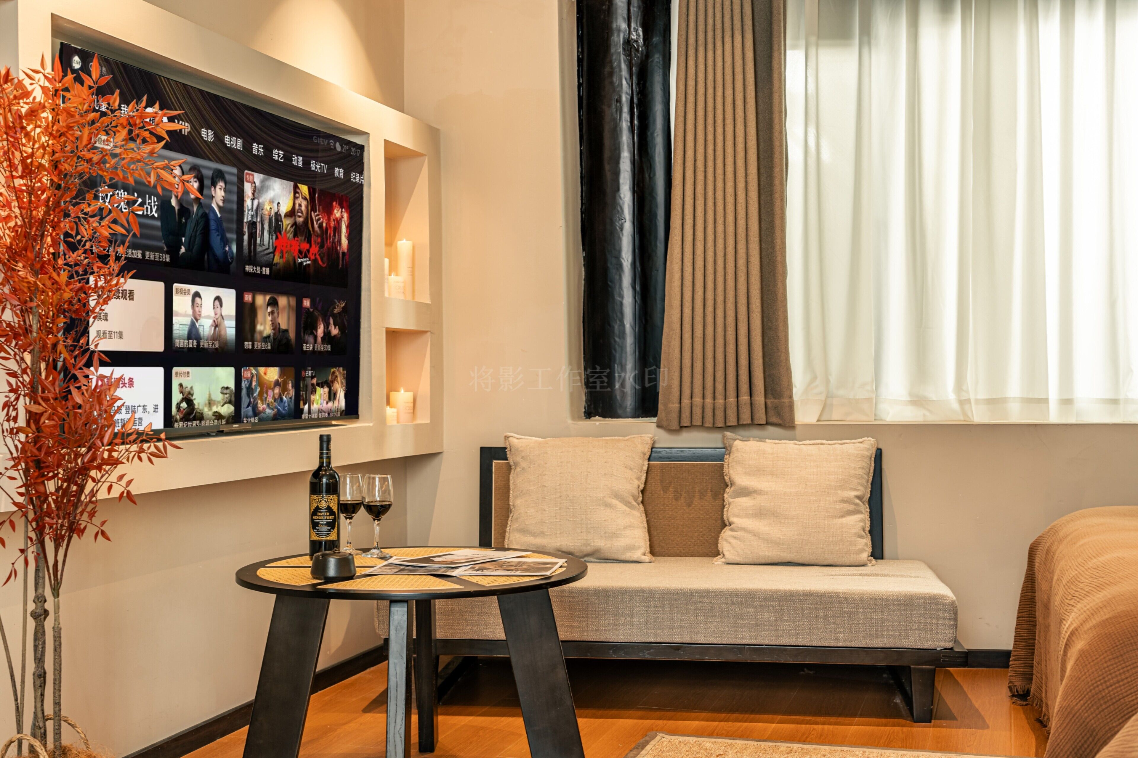 Quarto de hotel moderno com sofá, uma pequena mesa redonda com garrafa e copos, televisão presa à parede e janela com cortinas.