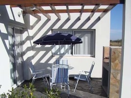 Um alpendre coberto com mesa e cadeiras, um guarda-sol azul e vista para a paisagem.