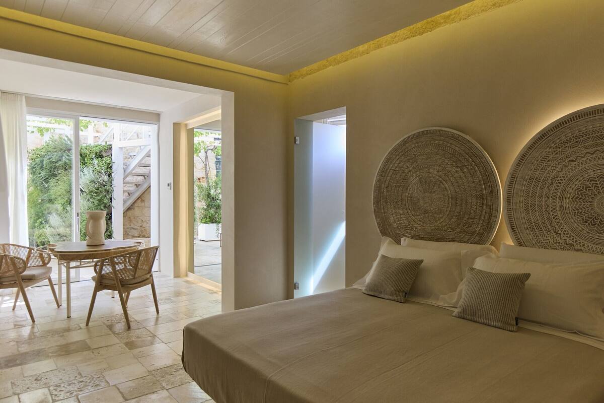 Quarto com cama, dois objetos decorativos na parede, área de refeições com mesa e cadeiras, e vista para uma escada que leva para o exterior.