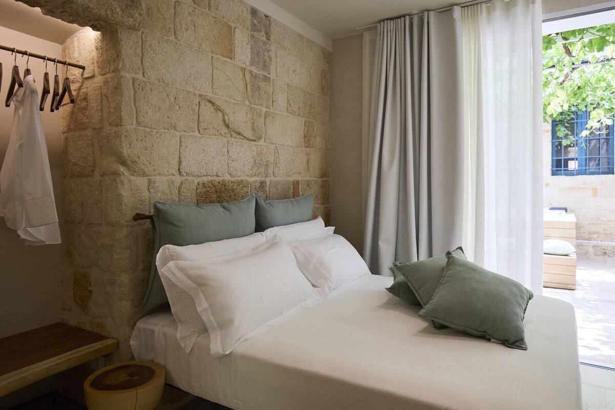 Quarto com parede de pedra, cama com almofadas brancas e verdes, um banco de madeira e uma janela com cortinas.