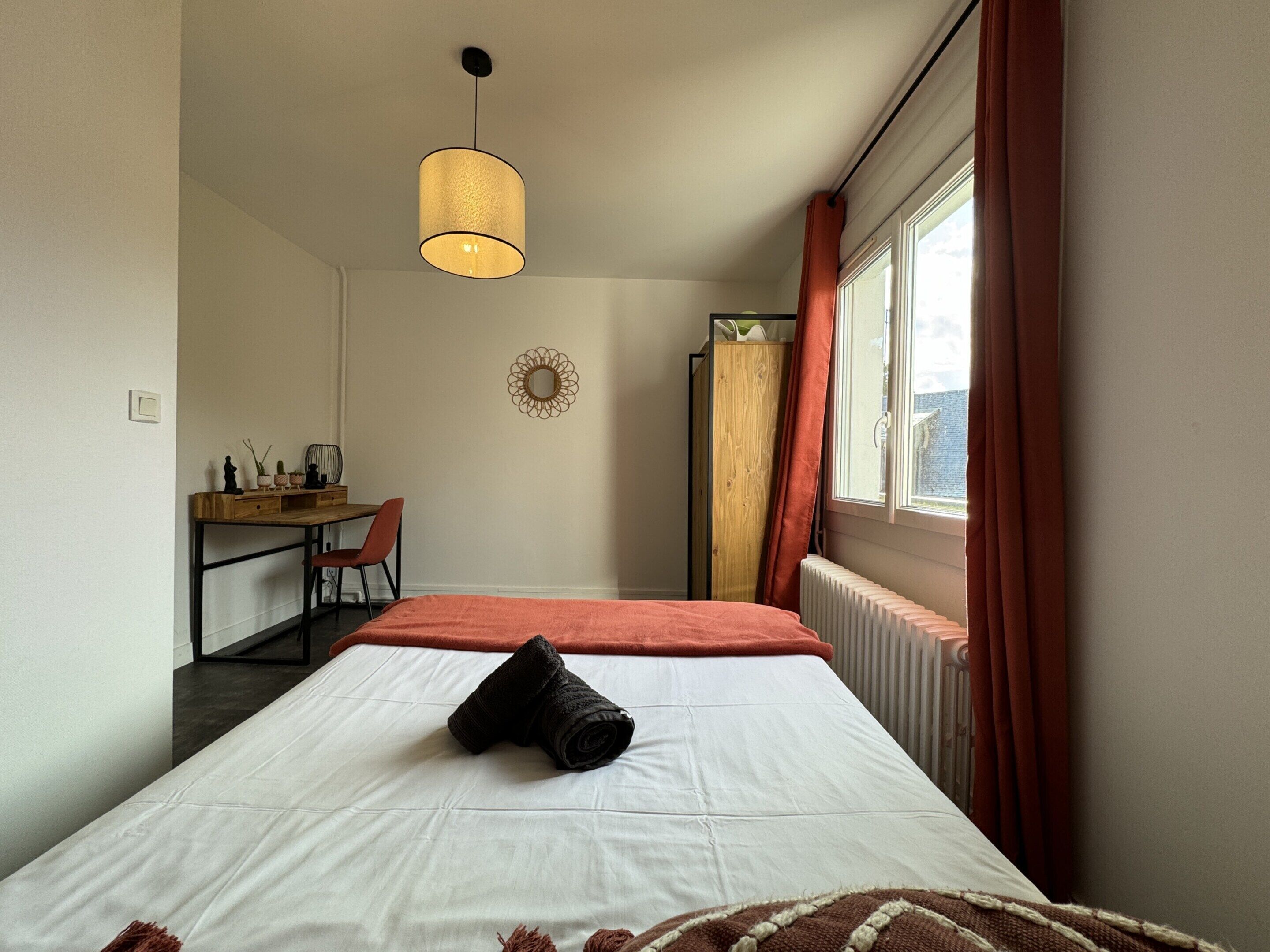 Quarto com cama, escrivaninha, cadeira, luminária pendente e janela com cortinas.
