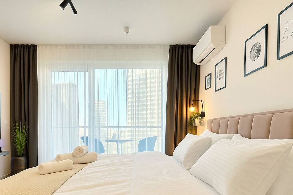 Quarto de hotel com uma cama grande, uma mesa de cabeceira com abajur, uma janela com cortinas e vista para prédios.