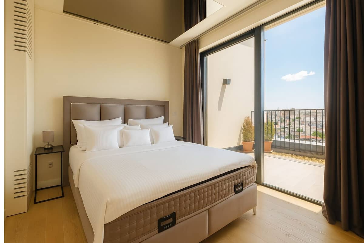Quarto de hotel com uma cama grande, uma mesa de cabeceira, janela com vista para a cidade e varanda com plantas em vasos.