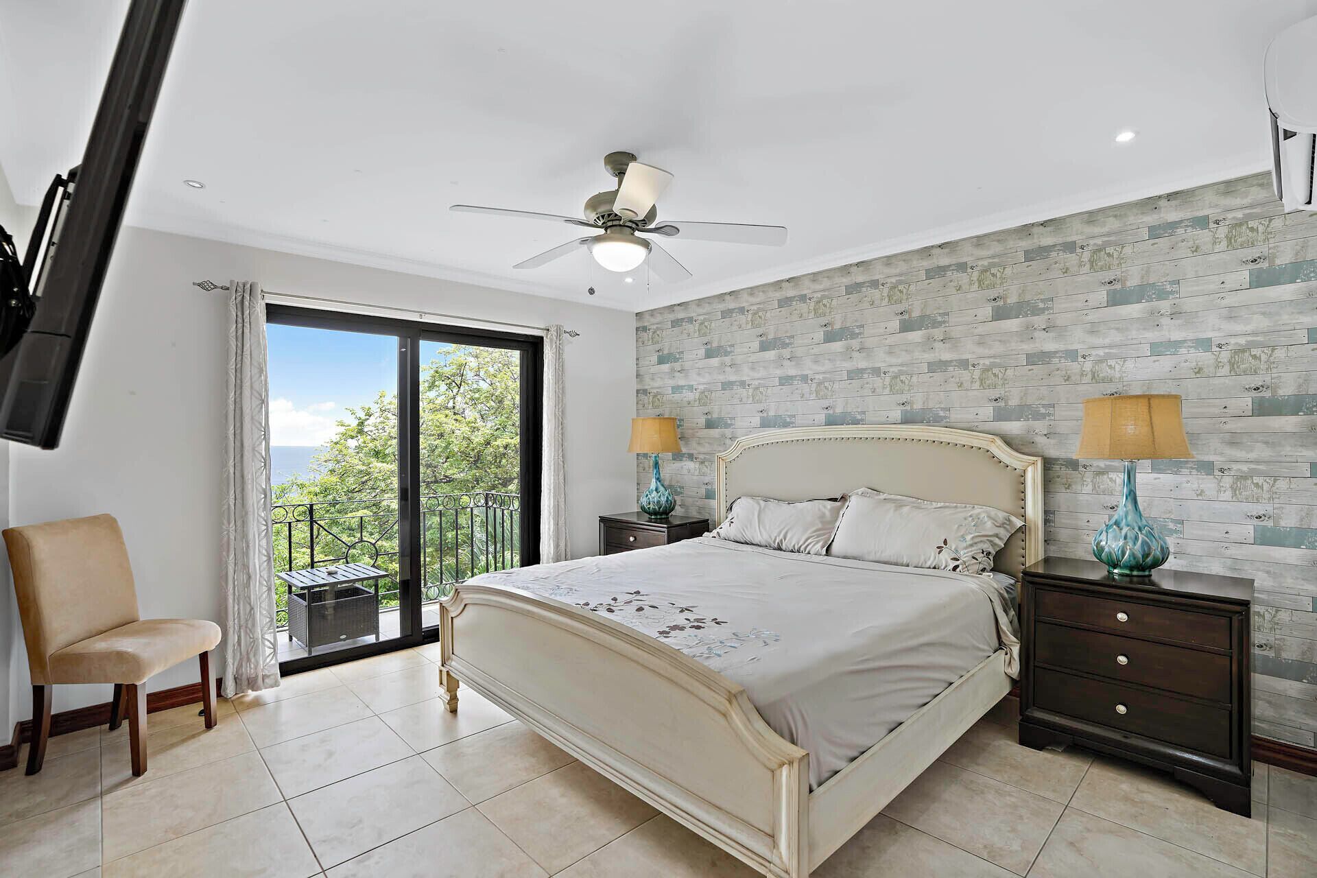 Quarto com cama, duas mesas de cabeceira, uma cadeira, ventilador de teto e varanda com vista.