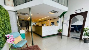 Um hall de hotel com uma mesa de recepção, uma área de estar com uma cadeira azul e um tapete marrom, e uma parede verde com uma planta.