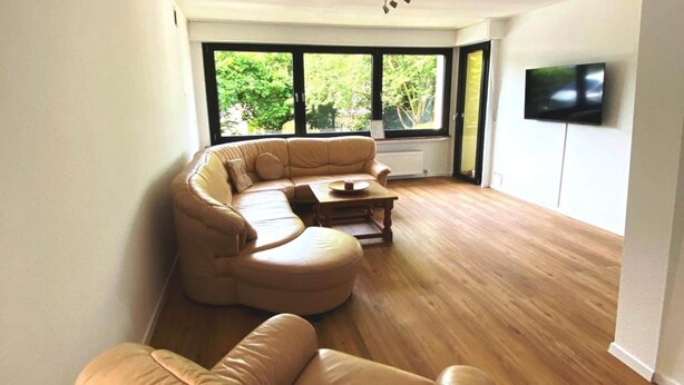 Uma sala de estar moderna com um sofá de couro, uma mesa de centro de madeira e uma televisão fixada na parede.