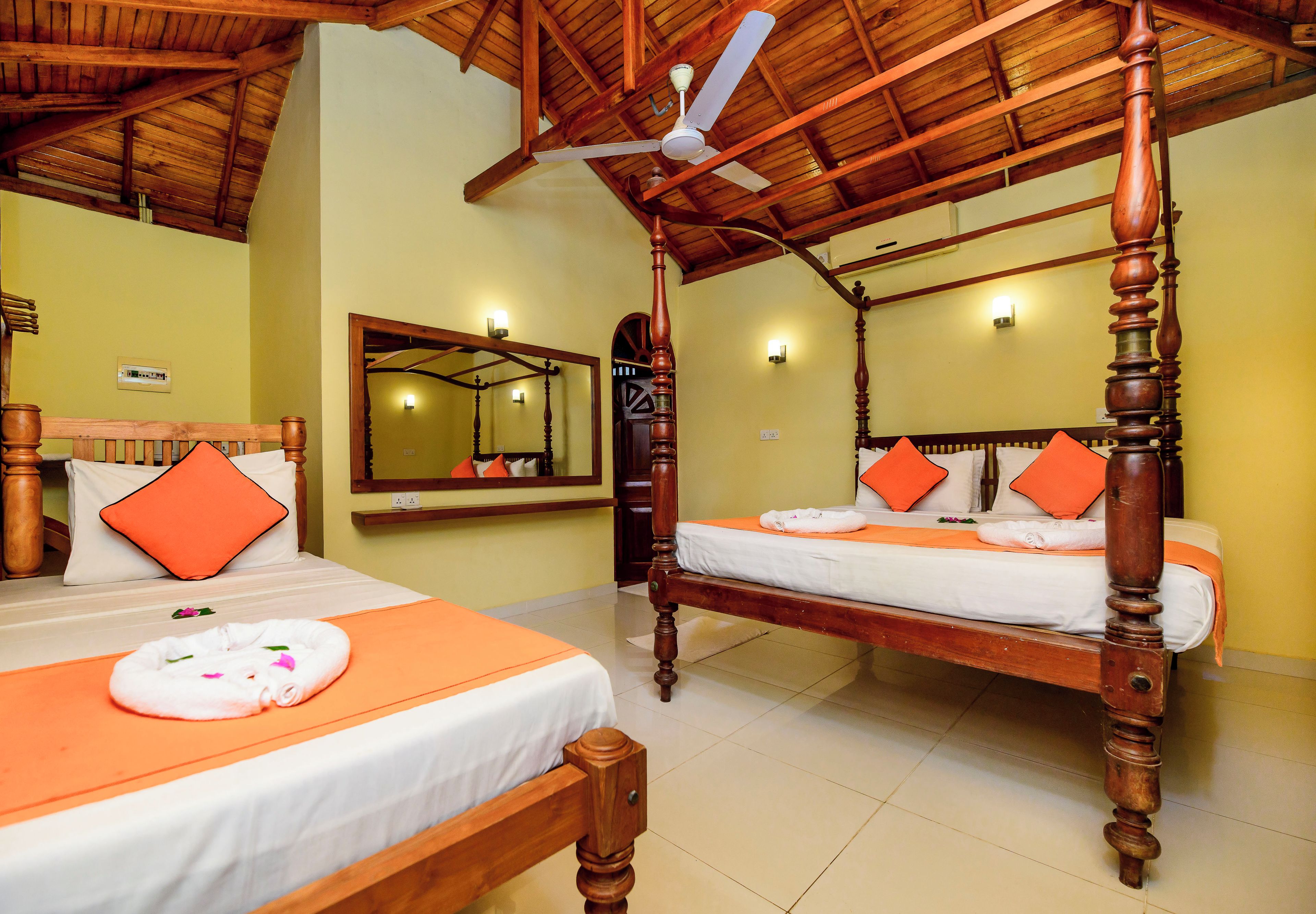 Quarto de hotel com duas camas de madeira, um ventilador de teto, um espelho e uma luminária na parede.
