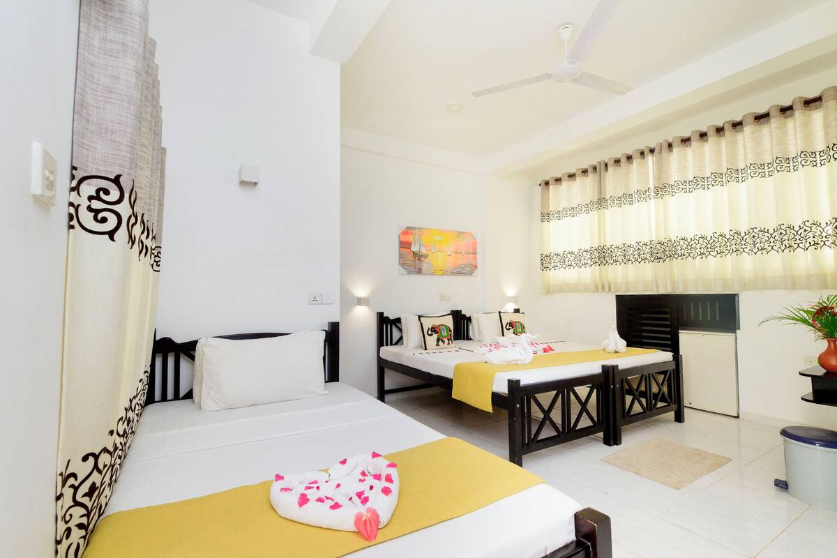 Quarto de hotel com duas camas, uma cozinha compacta, um ventilador de teto e uma janela com cortinas estampadas.