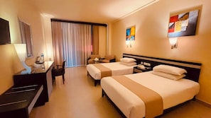 Quarto de hotel com duas camas, uma escrivaninha, uma cadeira, uma mesinha e uma janela com cortinas.