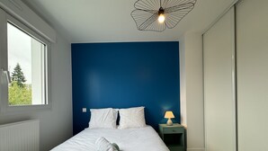 Quarto com cama grande, roupa de cama branca, parede azul de destaque, abajur e vista pela janela.