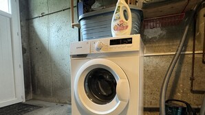 Máquina de lavar roupa com carga frontal numa lavanderia com chão de betão, um aspirador e uma garrafa de detergente em cima.