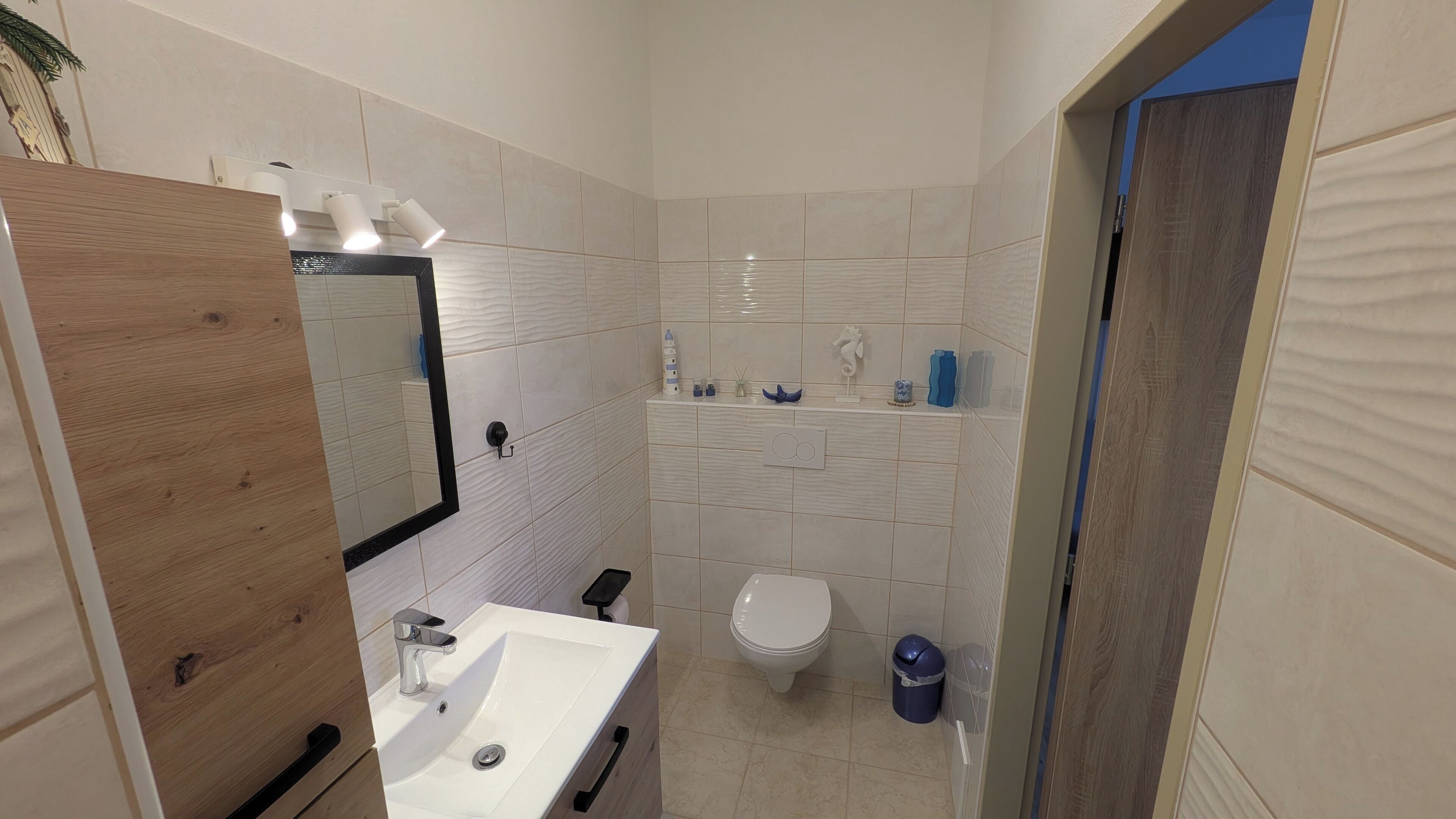 Um banheiro com pia branca, espelho e vaso sanitário. Há prateleiras com produtos de higiene e uma lixeira.