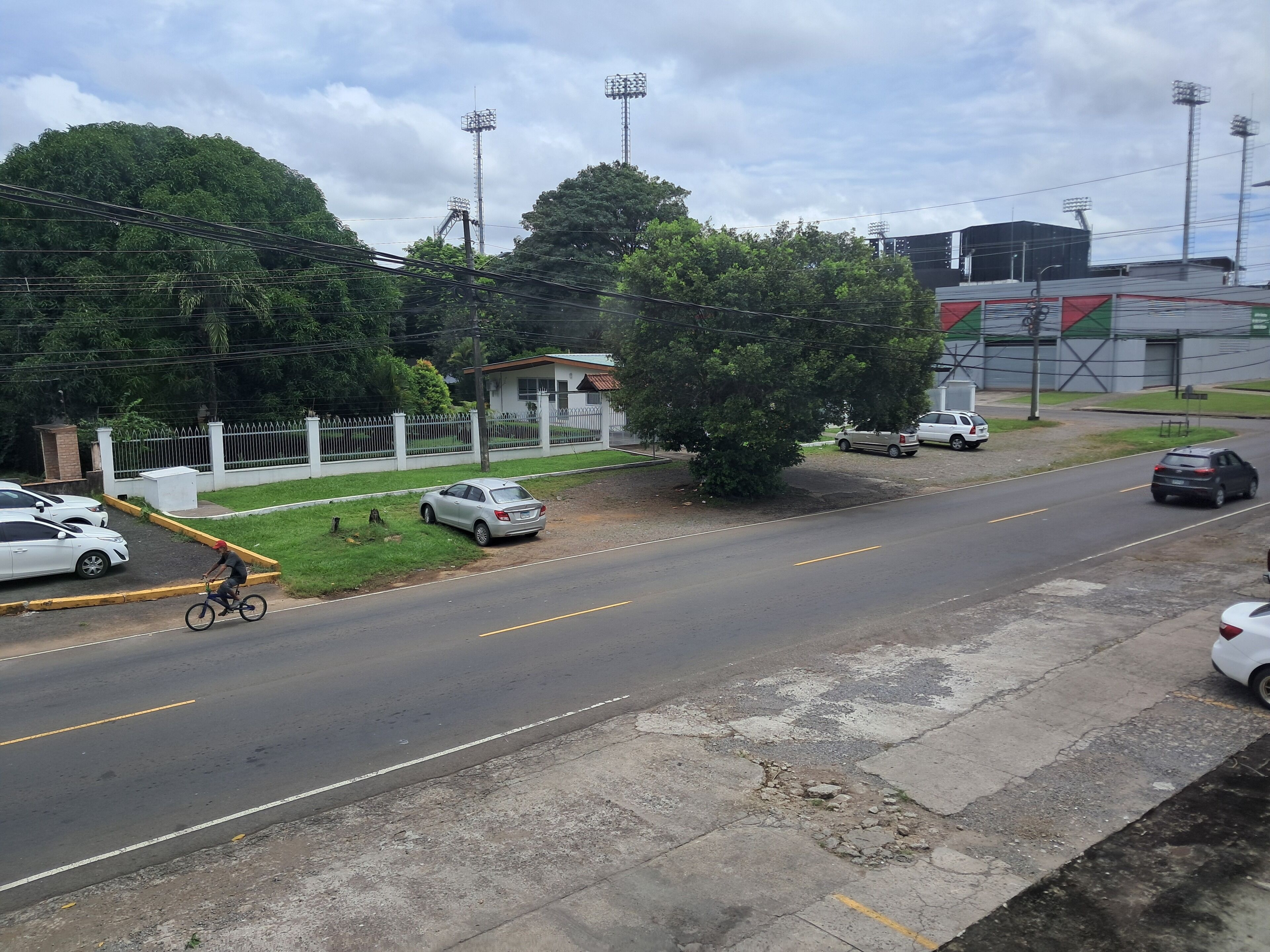 Uma estrada com carros e um ciclista. Árvores e uma área cercada com um edifício.