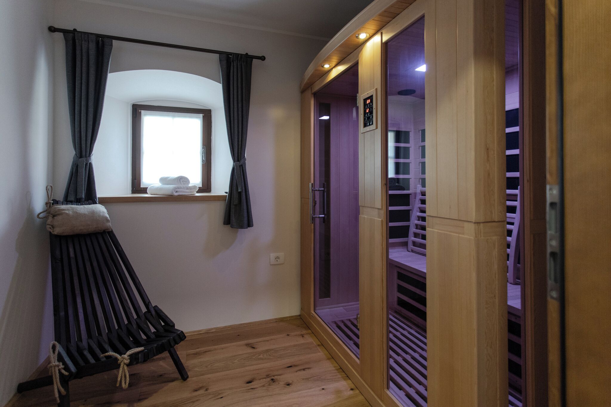 Um quarto com piso de madeira, uma poltrona de descanso preta, uma janela com cortinas e um guarda-roupa de madeira com espelho.