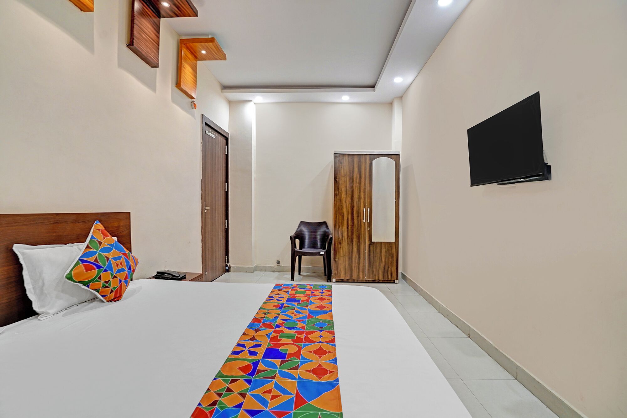 Quarto de hotel com cama, um tapete colorido, uma cadeira, guarda-roupa e televisão na parede.