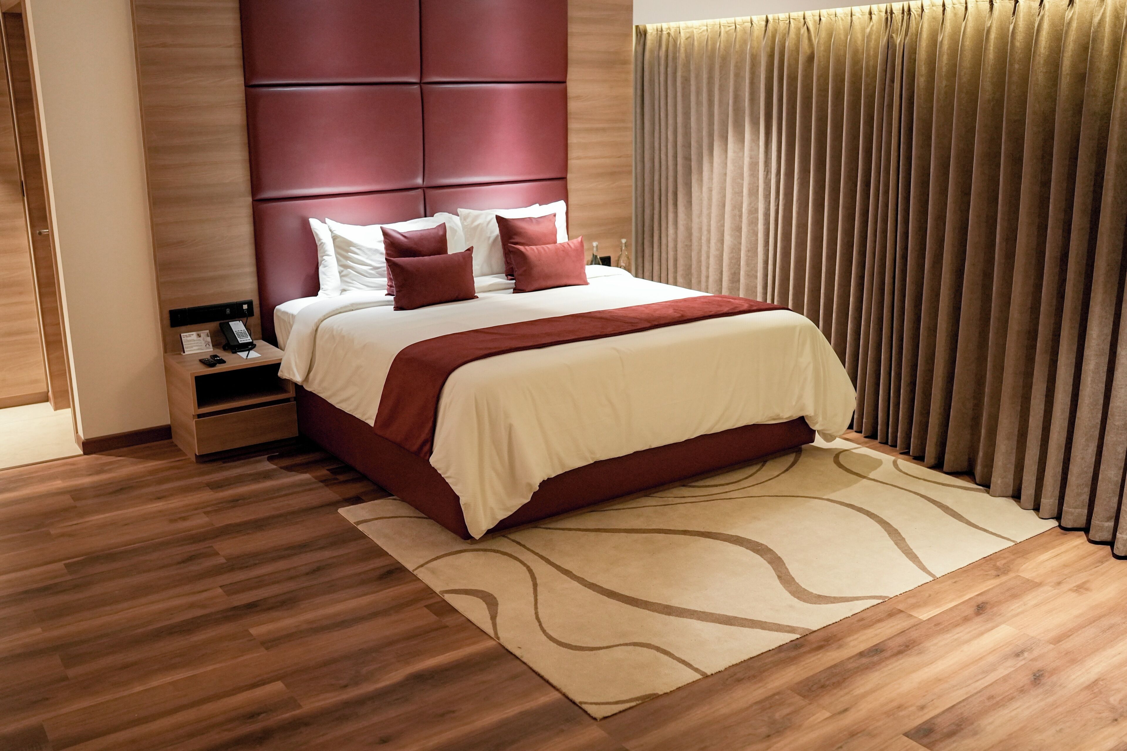 Quarto de hotel moderno com uma cama grande, cabeceira de madeira, mesa de cabeceira com telefone e abajur, e uma janela ampla com cortinas.