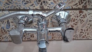 Uma torneira cromada com três maçanetas distintas, provavelmente para água quente, fria e possivelmente uma função economizadora, fixada em uma parede revestida de azulejos com padrão floral.