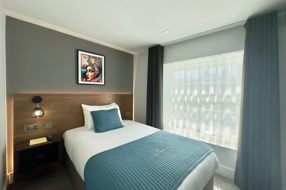 Quarto de hotel com cama, mesa de cabeceira, luminária na parede, obra de arte emoldurada e janela com cortinas.