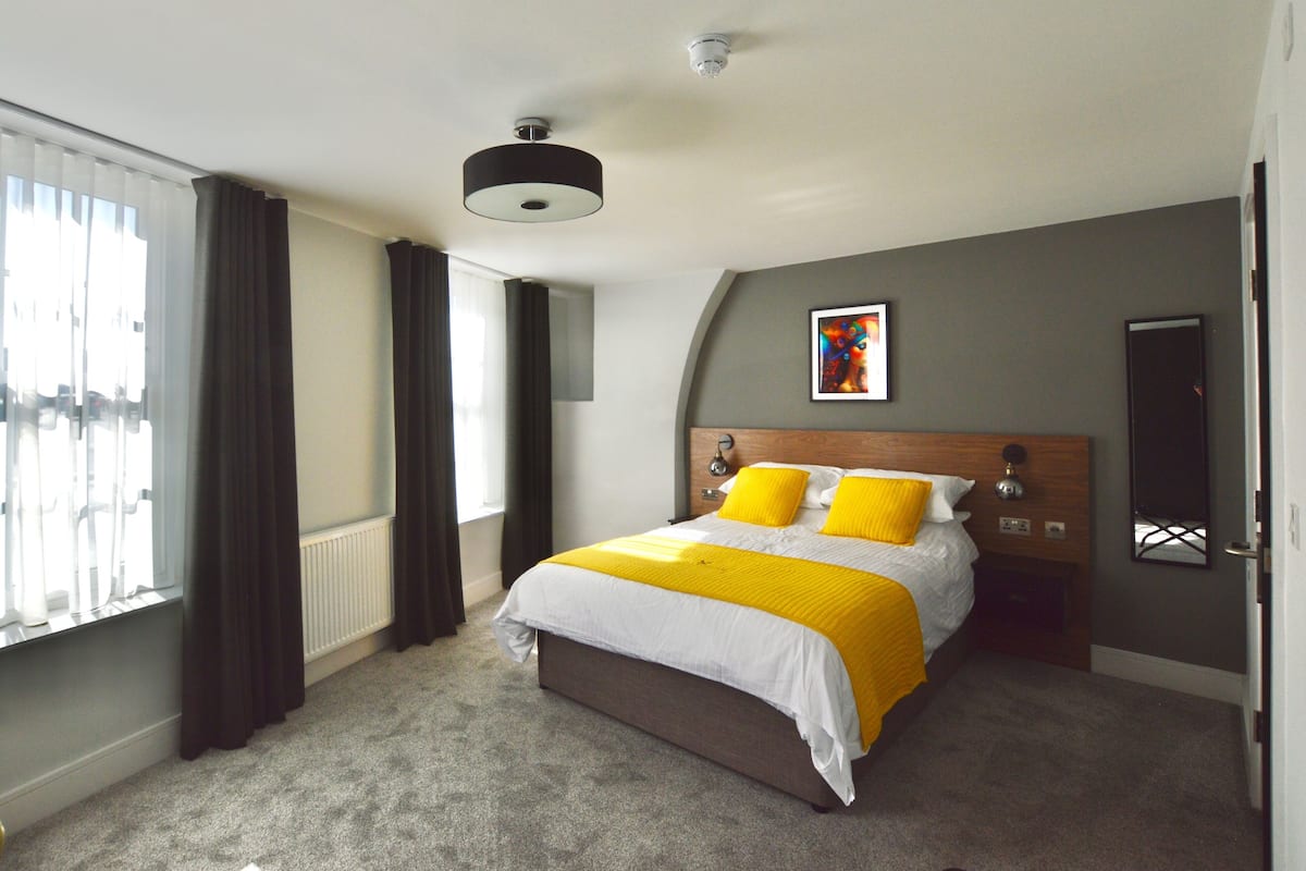 Quarto de hotel com uma cama grande, uma luminária no teto, uma janela com cortinas, um radiador e um quadro na parede.