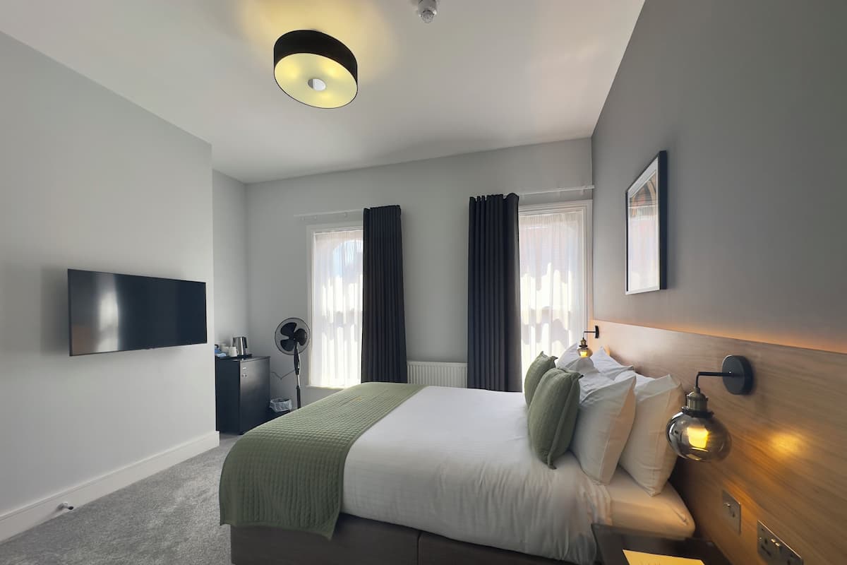 Quarto de hotel moderno com uma cama grande, televisão de tela plana, mesa de cabeceira com abajur e janela com cortinas.