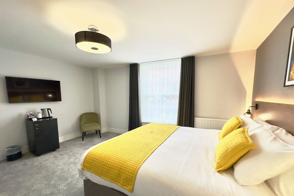 Quarto de hotel com uma cama, escrivaninha, cadeira, televisão e janela com cortinas.